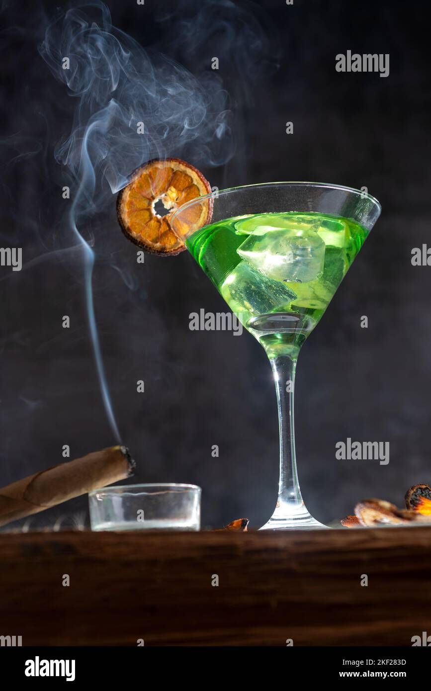 A closeup of green cocktail with lemon slice and cigar smoking on light ...