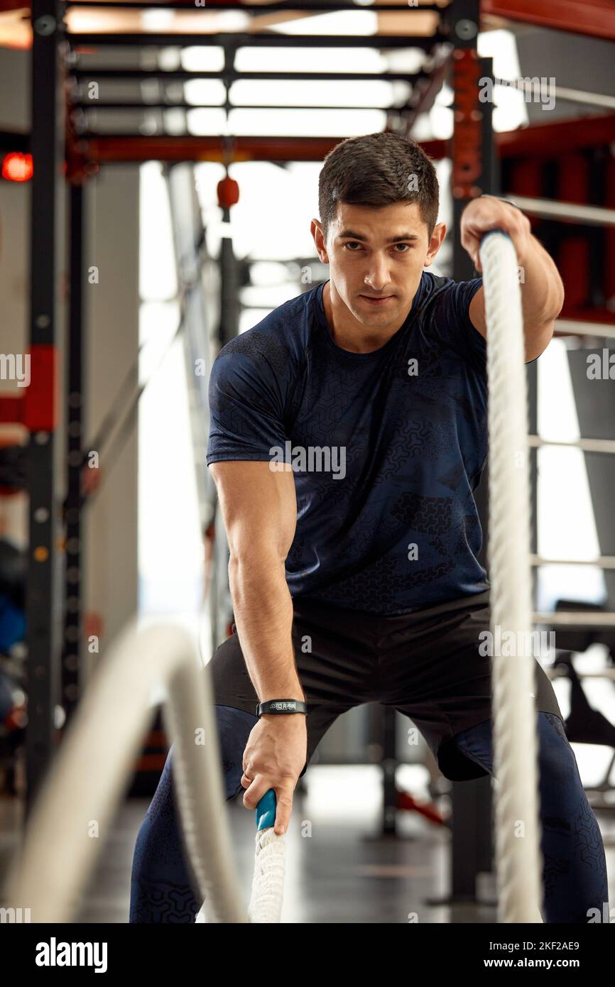 young handsome man in a black t-shirt is engaged with ropes in the gym ...