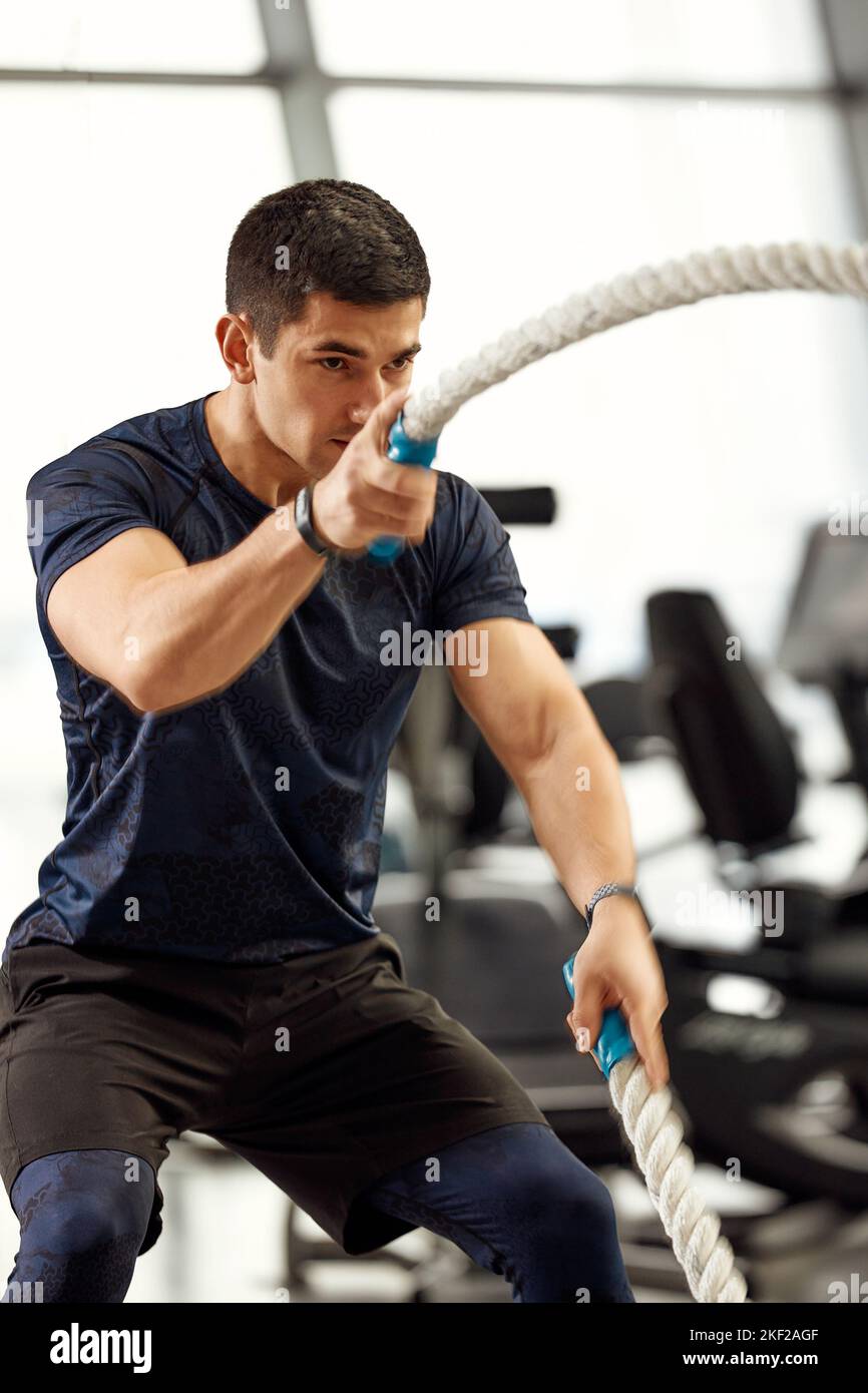 young handsome man in a black t-shirt is engaged with ropes in the gym ...