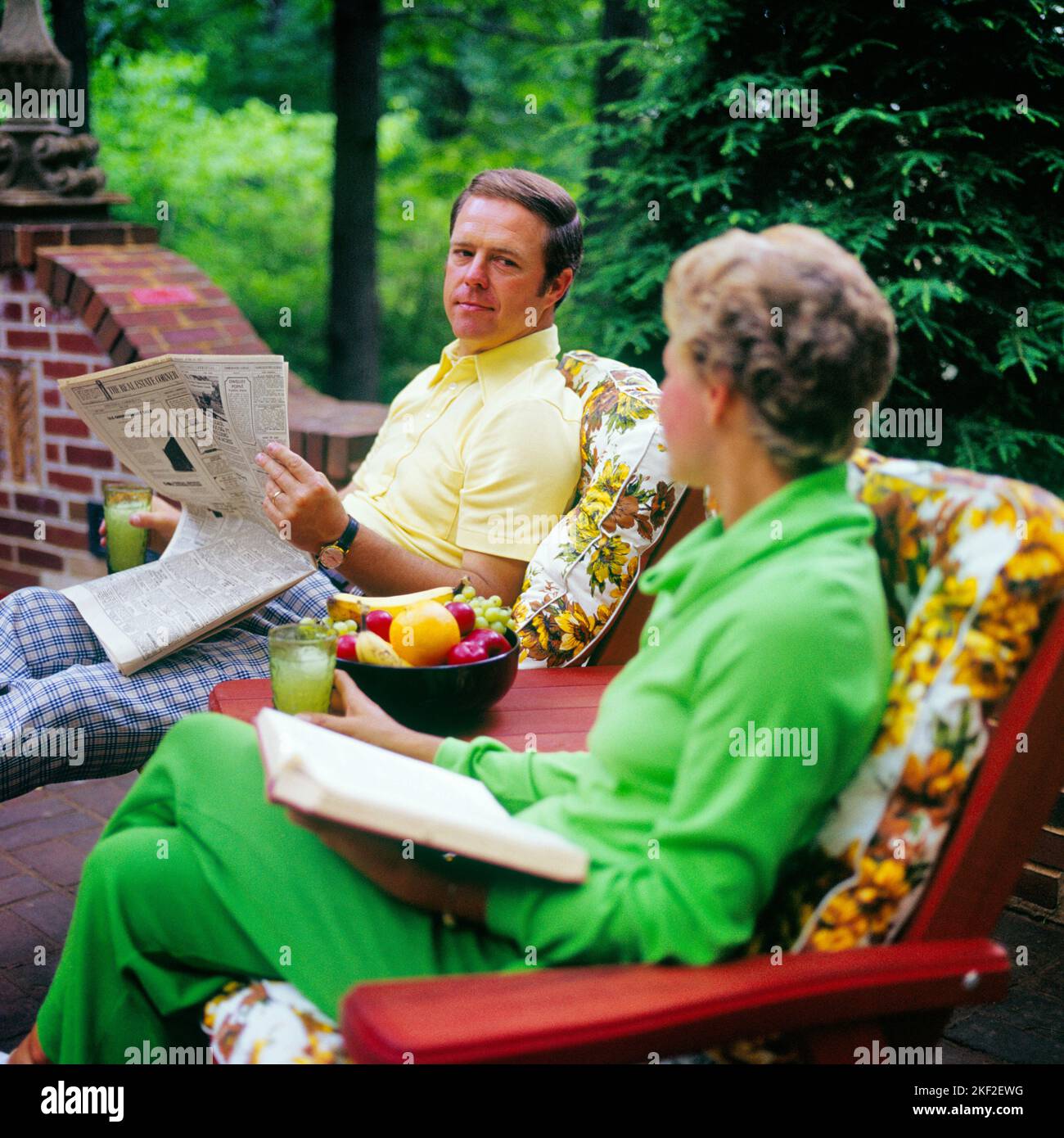 1970s COUPLE MAN AND WOMAN TALKING READING NEWSPAPER SITTING IN PATIO CHAIRS ON BACKYARD BRICK ...