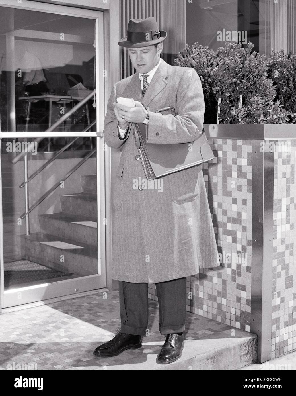 1950s SALESMAN WEARING TOP COAT & HAT WRITING NOTE IN PAD BY GLASS DOOR ...
