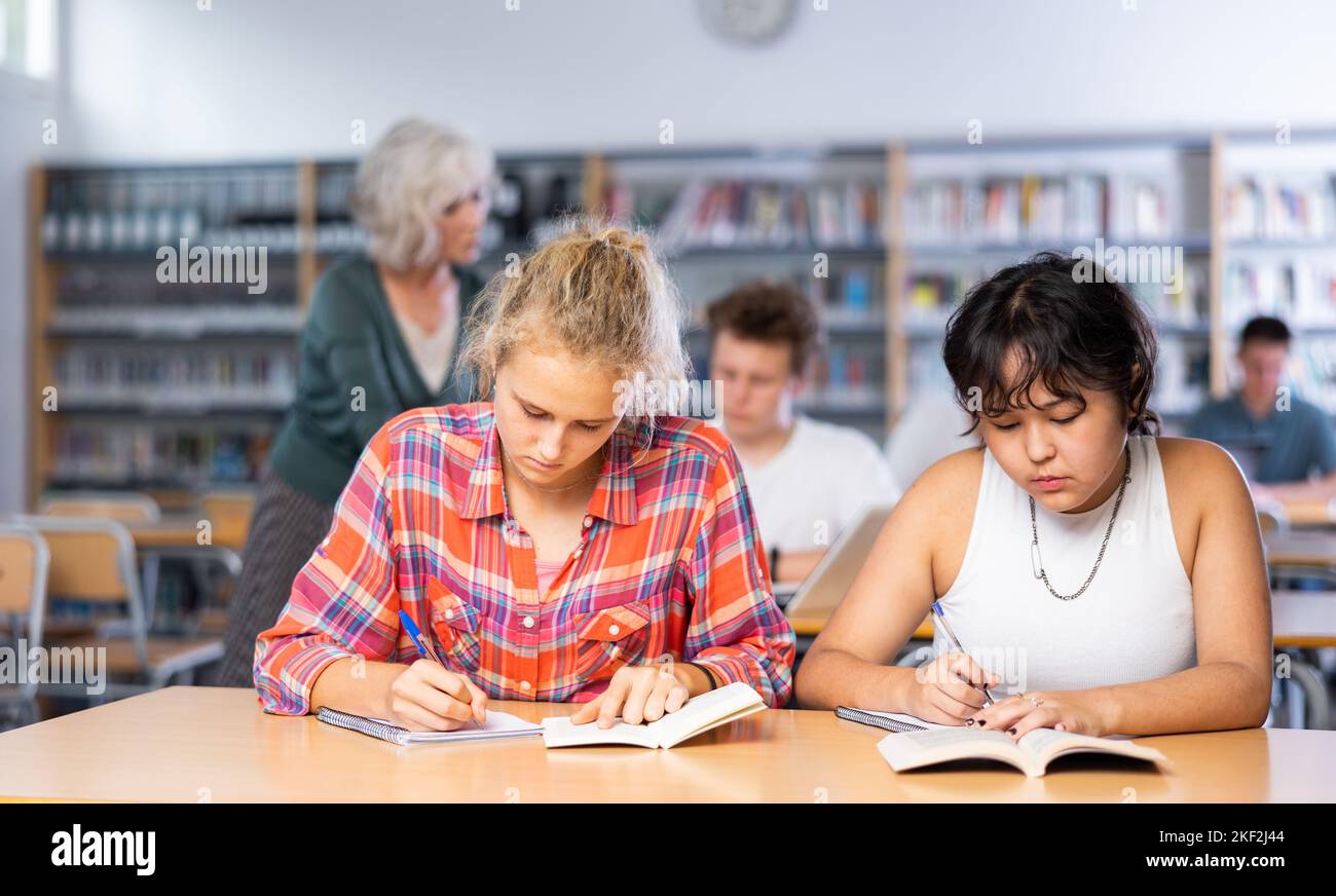 Two teenage friends sit at a desk in school library and carefully do ...