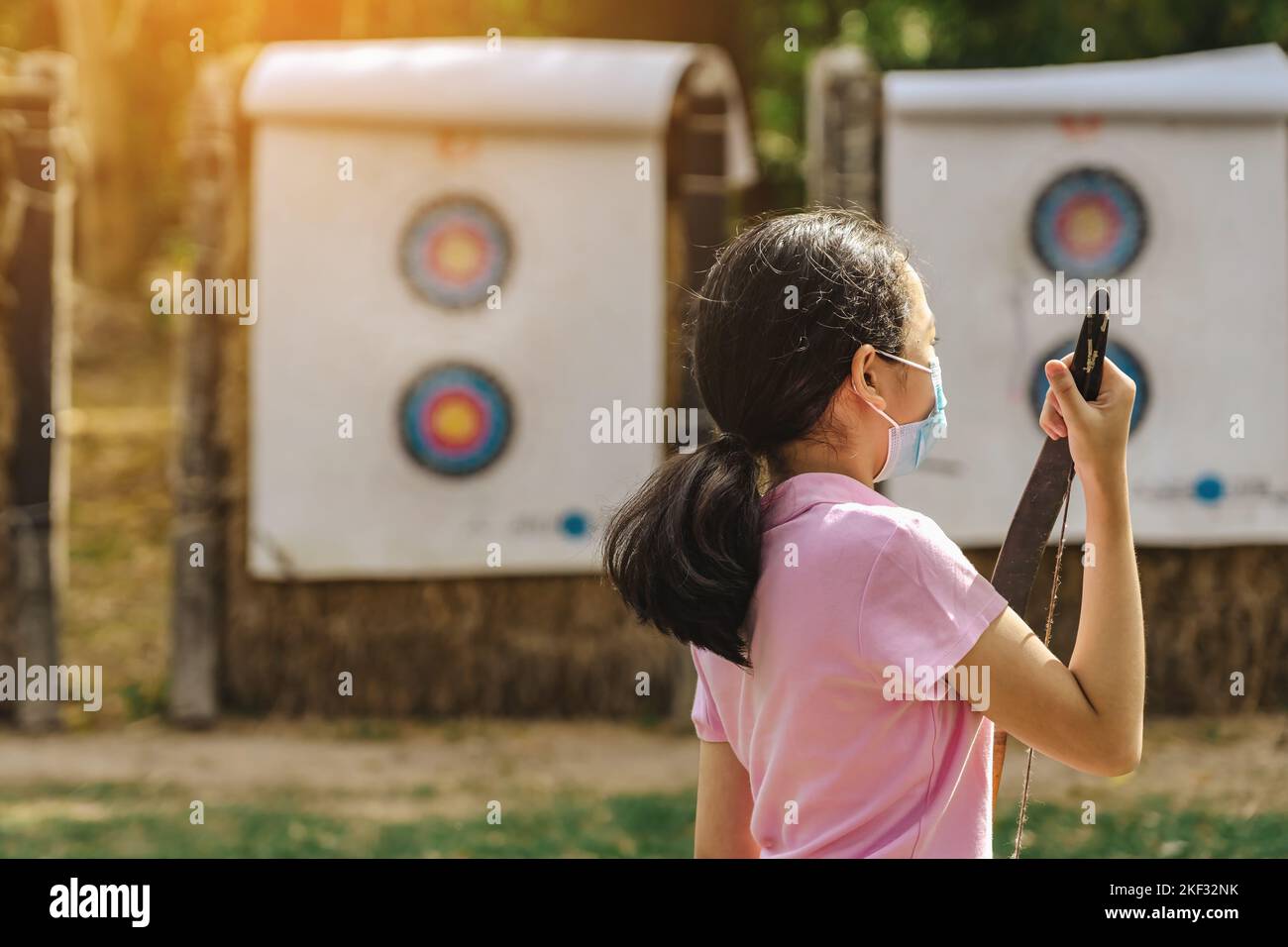 Back view of Asian girl wear face mask aims archery bow and arrow to ...