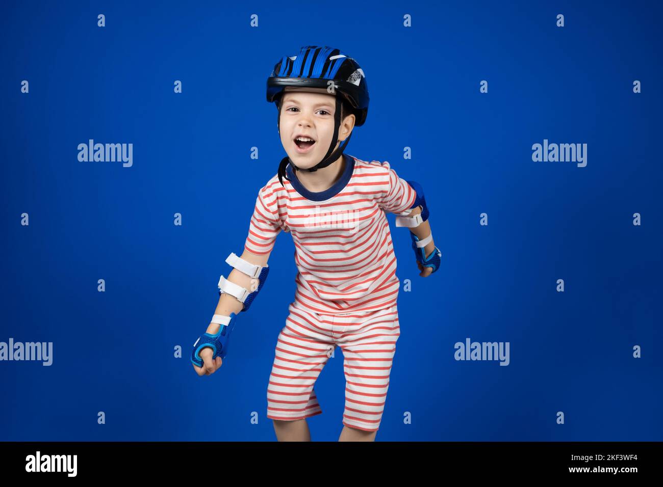 A happy boy in a protective helmet, elbow pads and gloves on a blue ...
