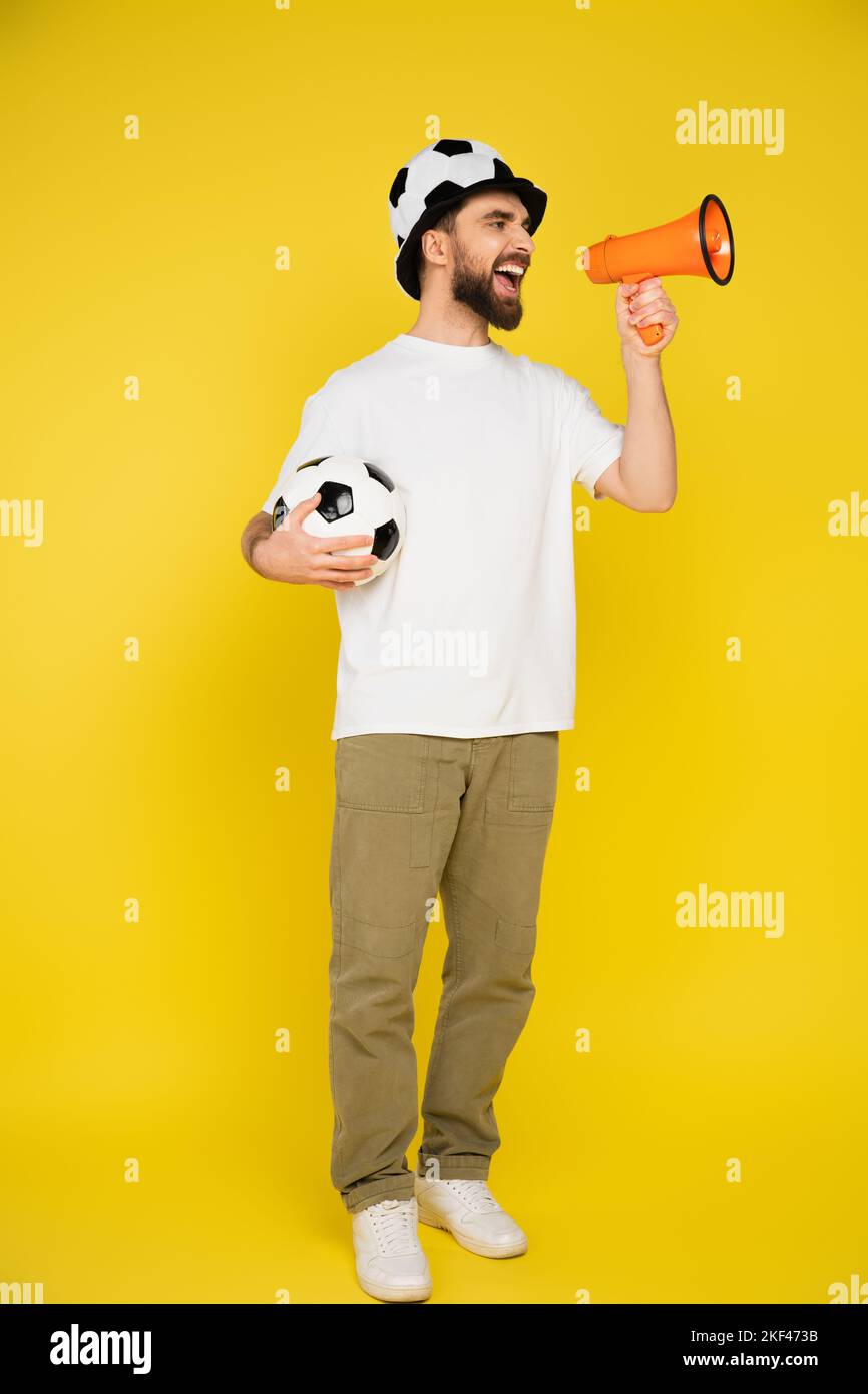 full length of sports fan in hat holding soccer ball and screaming in ...