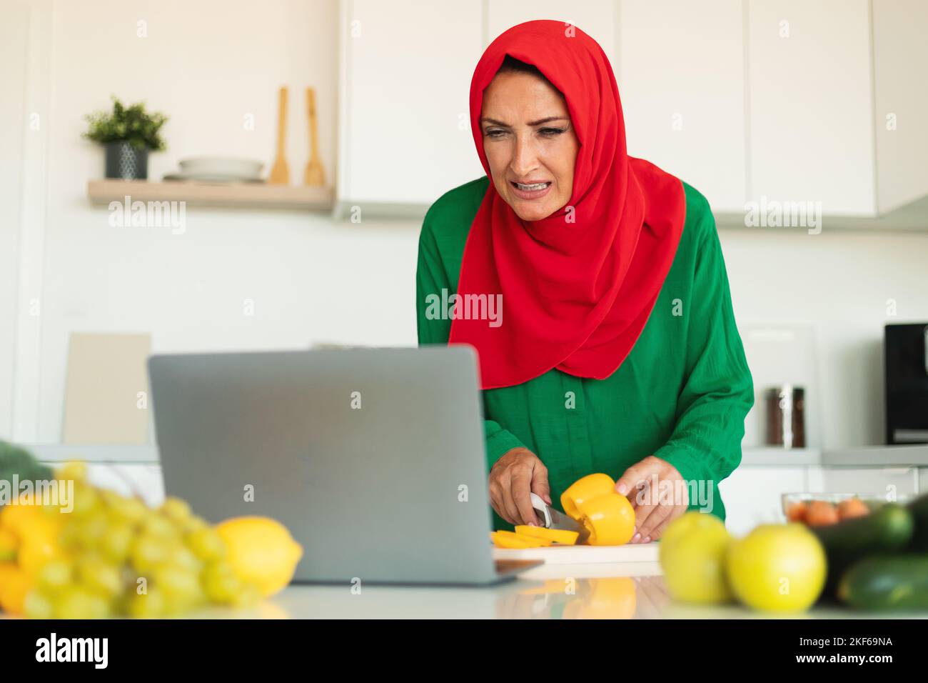 Mature islamic woman cooking, squinting eyes and looking at laptop ...