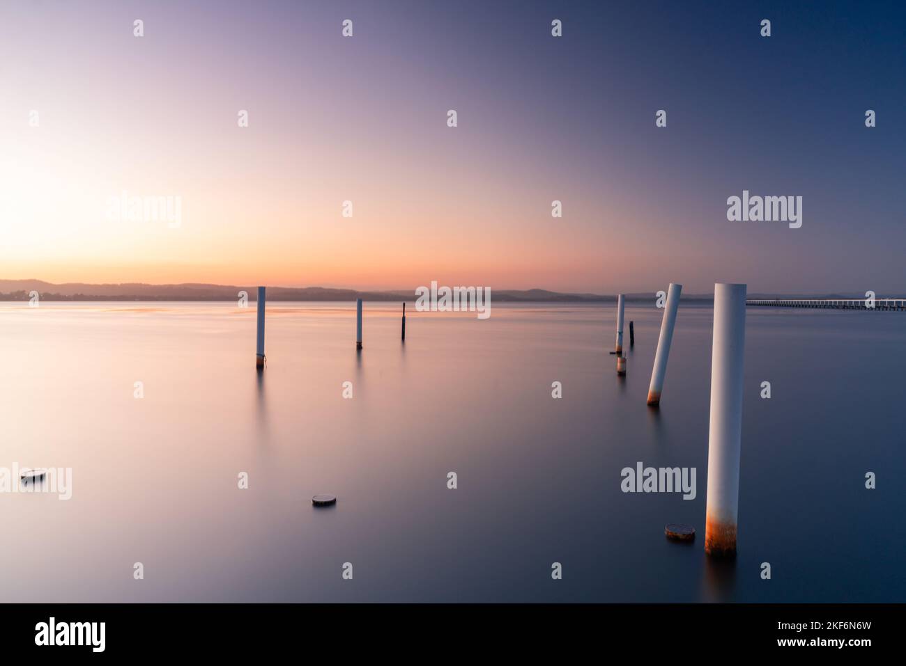 A scenic shot of a gradient sky reflecting on the calm water with posts ...