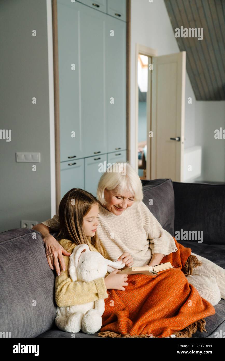 White girl reading book with her grandmother while siting on couch at ...