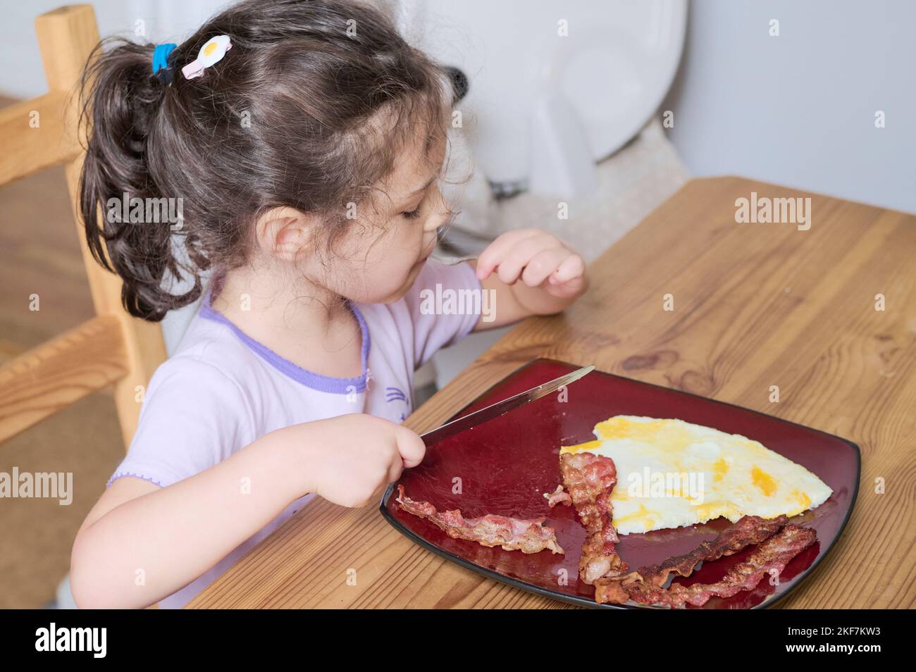 Young girl eating eggs and bacon with knife and fork for the first time ...
