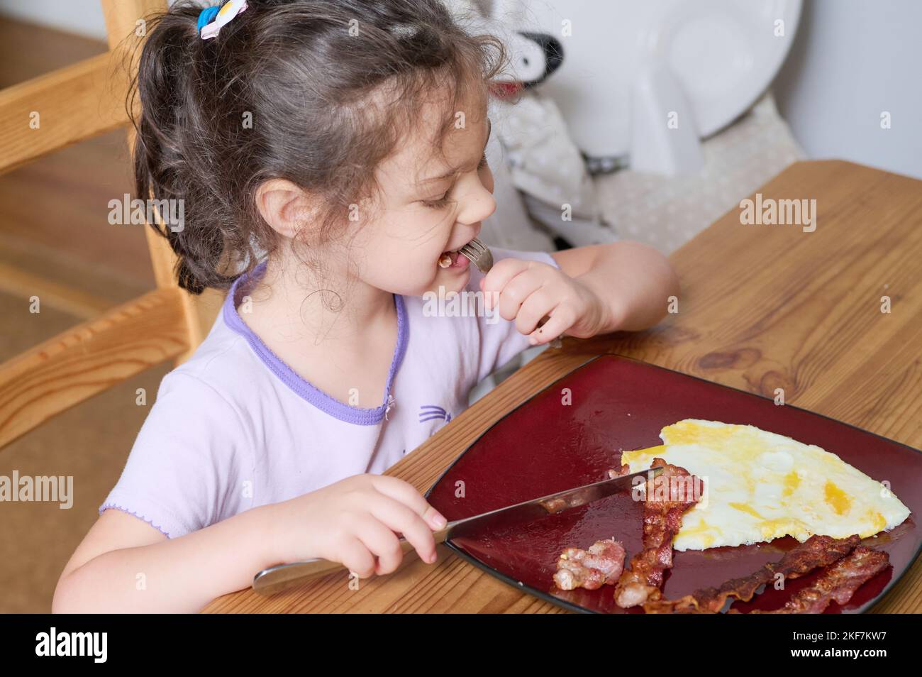 Young girl eating eggs and bacon with knife and fork for the first time ...
