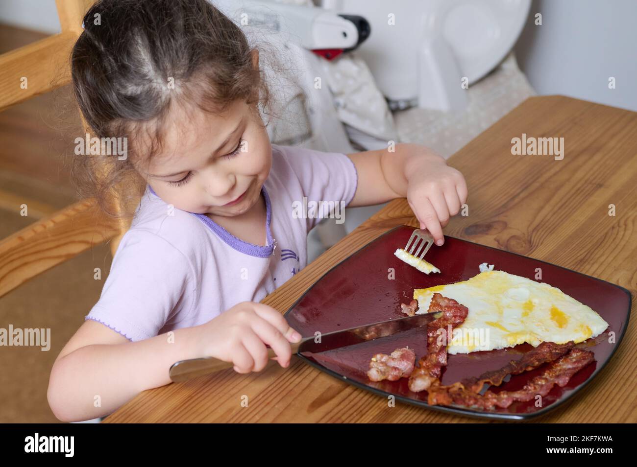 Young girl eating eggs and bacon with knife and fork for the first time ...