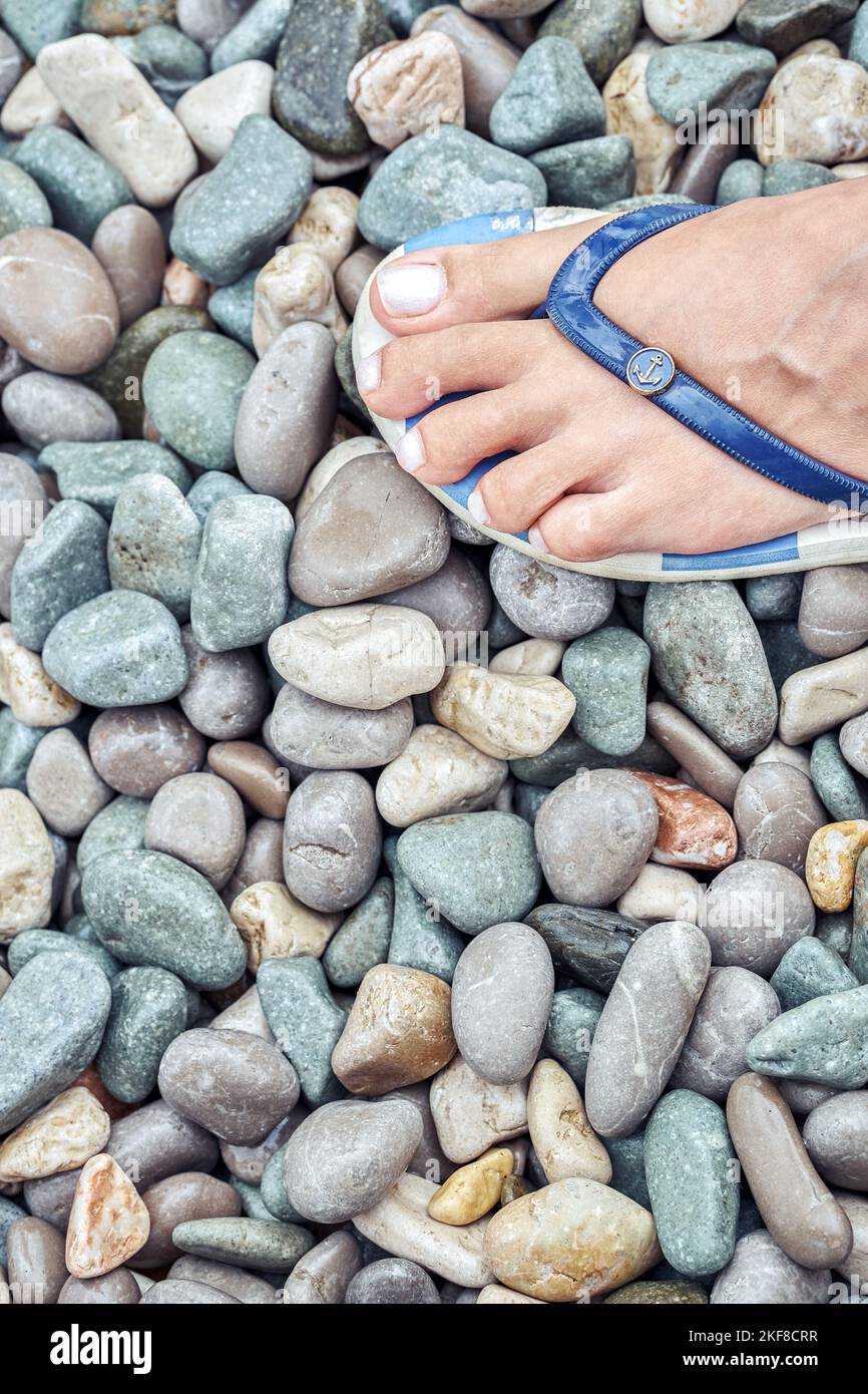 Tourist foot in bright blue flip-flops stands on pebble beach near sea ...