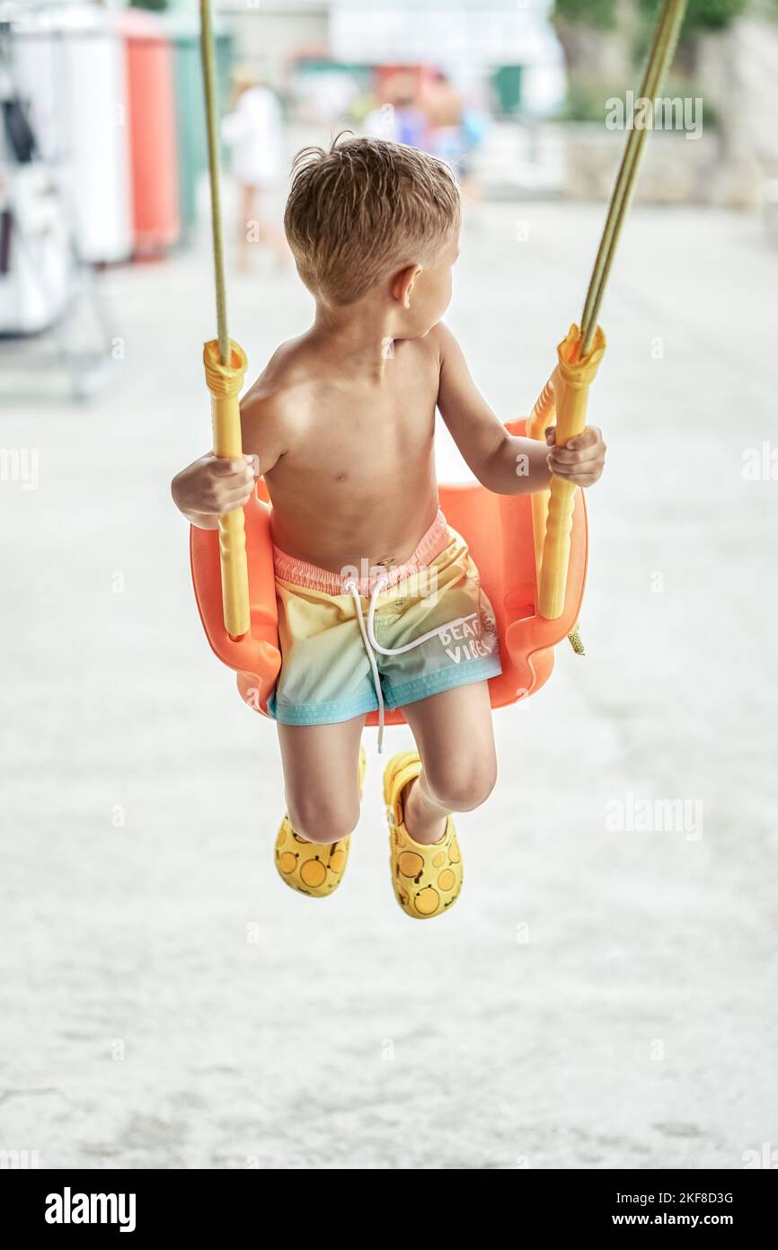 Small blonde boy swinging on an orange swing and looks back. Small child swings on hotel ...