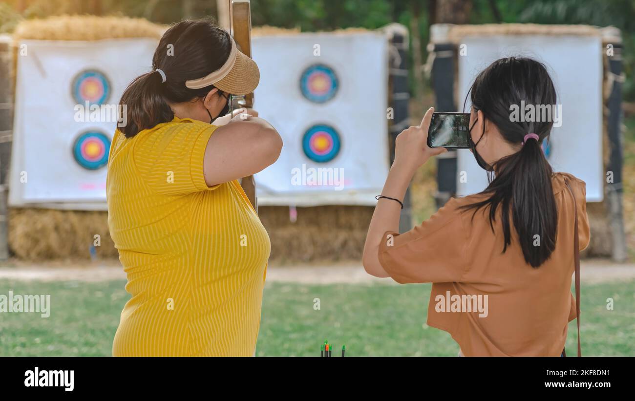 Back view of Asian woman wear visor with face mask aims archery bow and ...