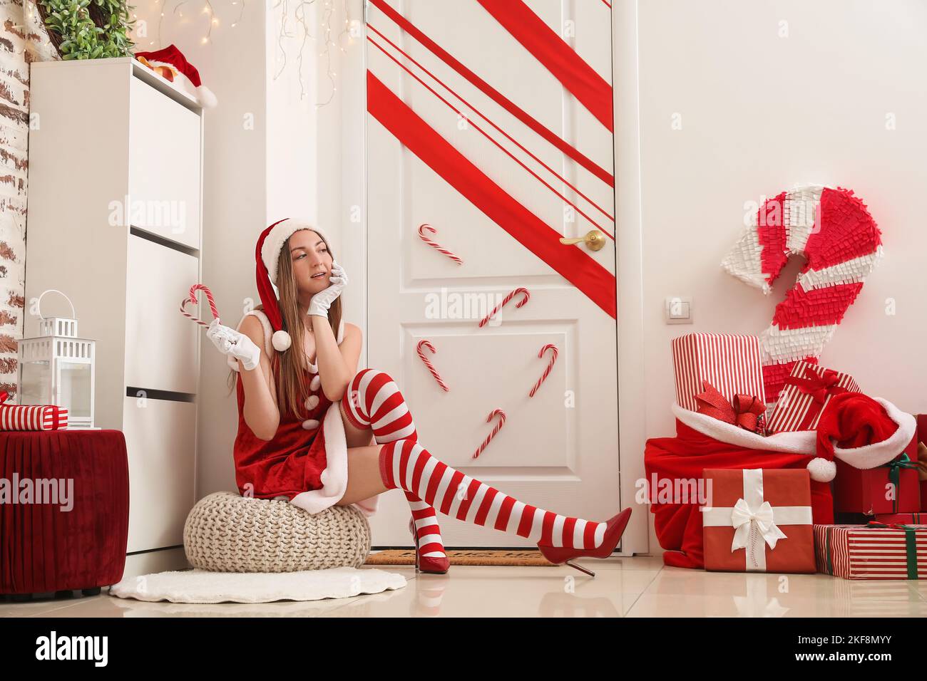 Young woman in Santa costume with candy canes sitting near white door ...