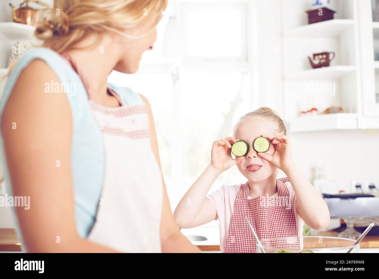 Having fun in the kitchen. Cute little girl playfully holding cucumber ...