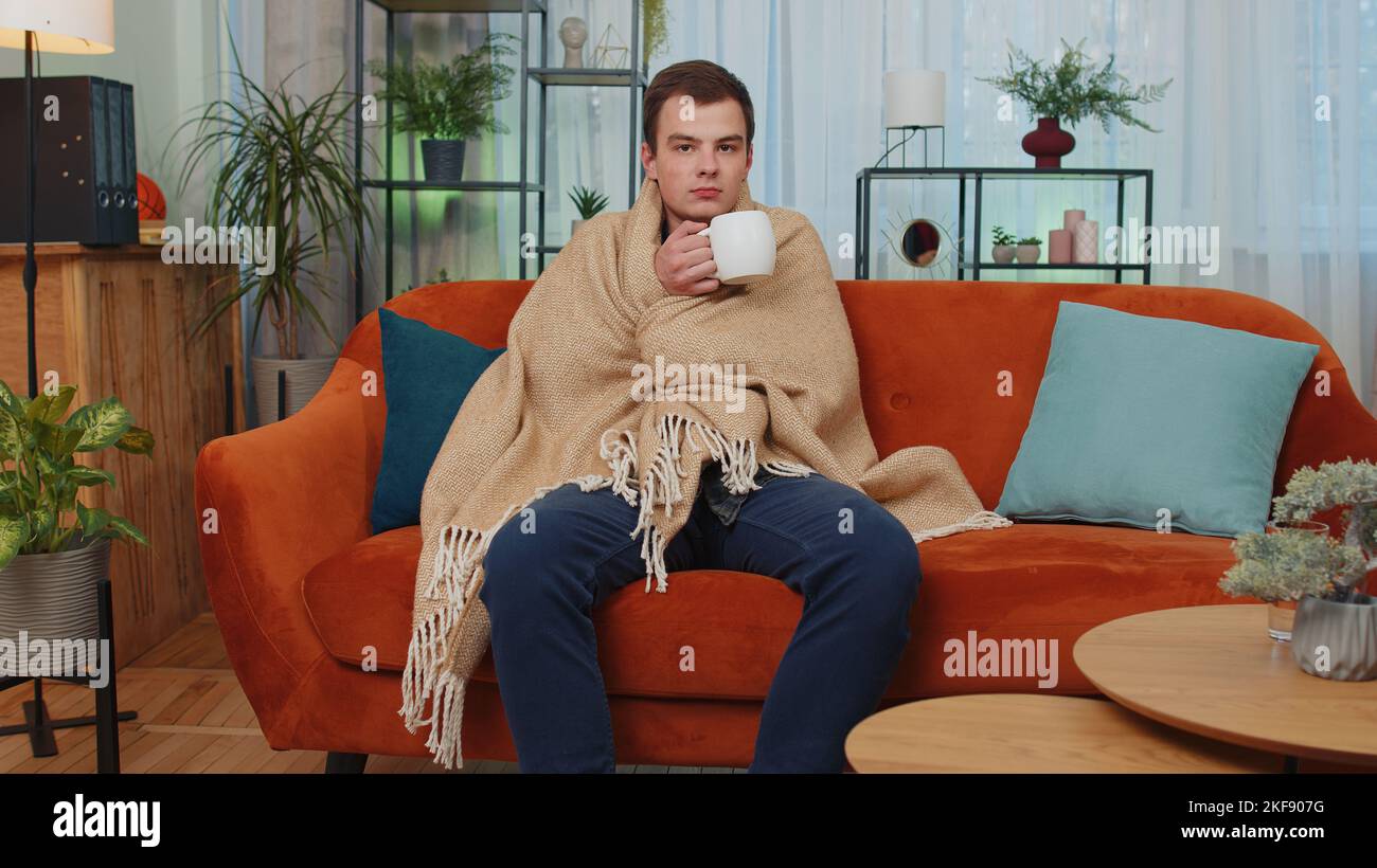 Sick man student wrapped in plaid shivering from cold on sofa drinking ...