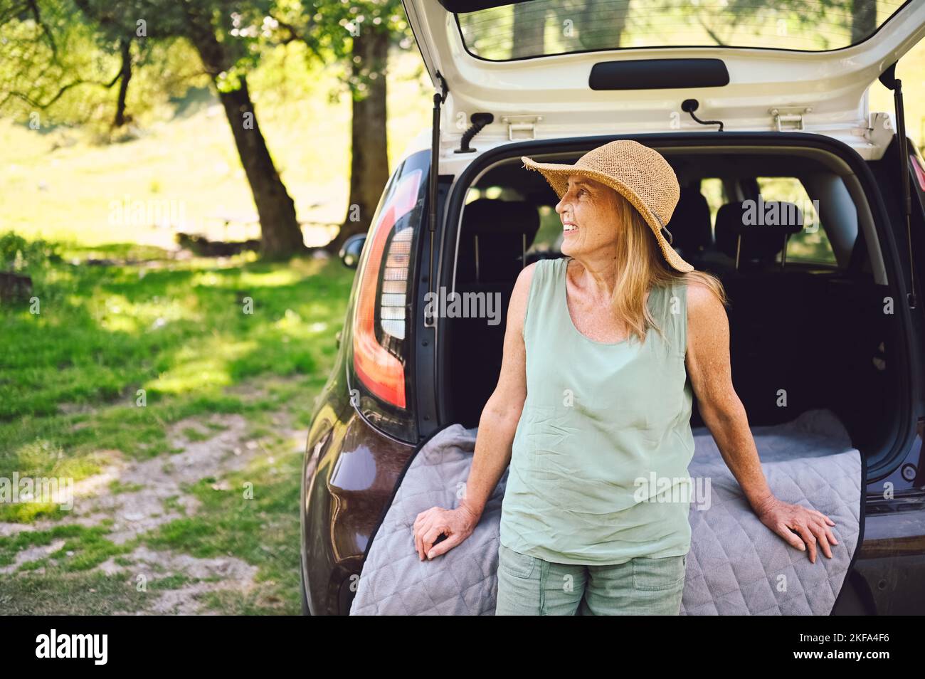 Happy senior woman in straw hat sitting in car trunk or boot outdoors ...