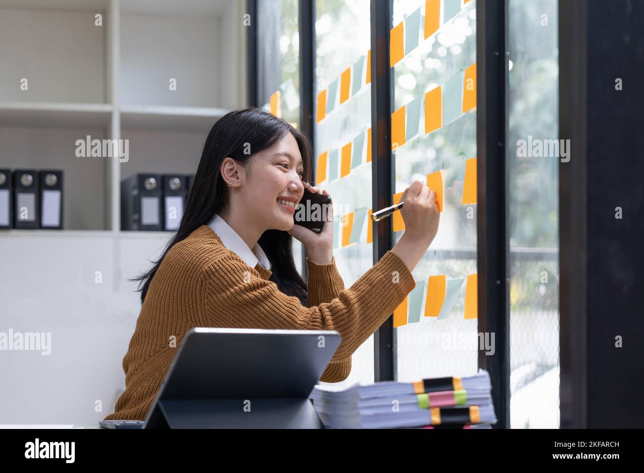 Smiling young asian woman writing on sticky notes on window in creative ...