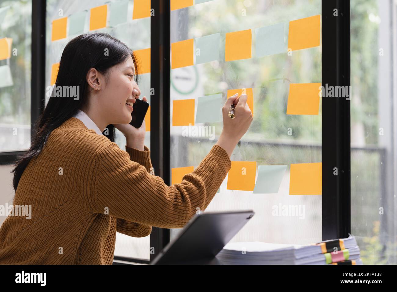 Smiling young asian woman writing on sticky notes on window in creative ...