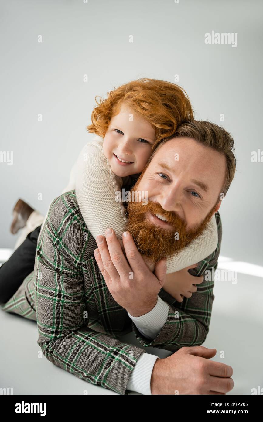 Red haired boy hugging father while lying on grey background with ...