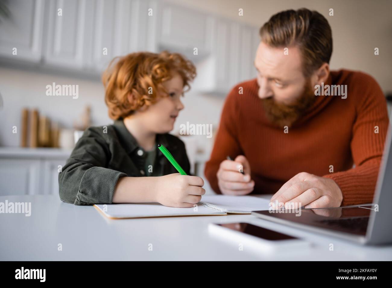 bearded man talking to redhead son doing homework near blurred laptop ...