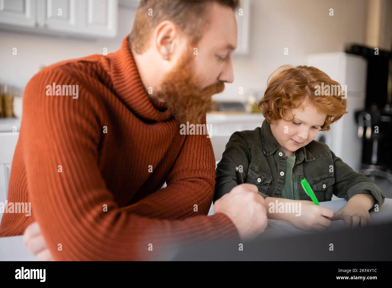 bearded man looking at redhead son writing in notebook while doing ...