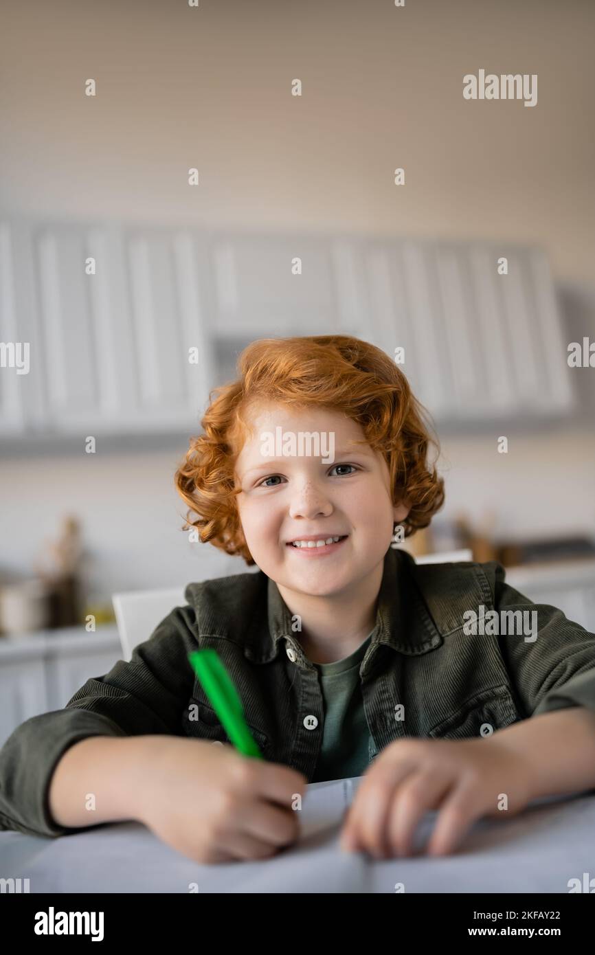 happy redhead kid with pen looking at camera while doing homework on ...