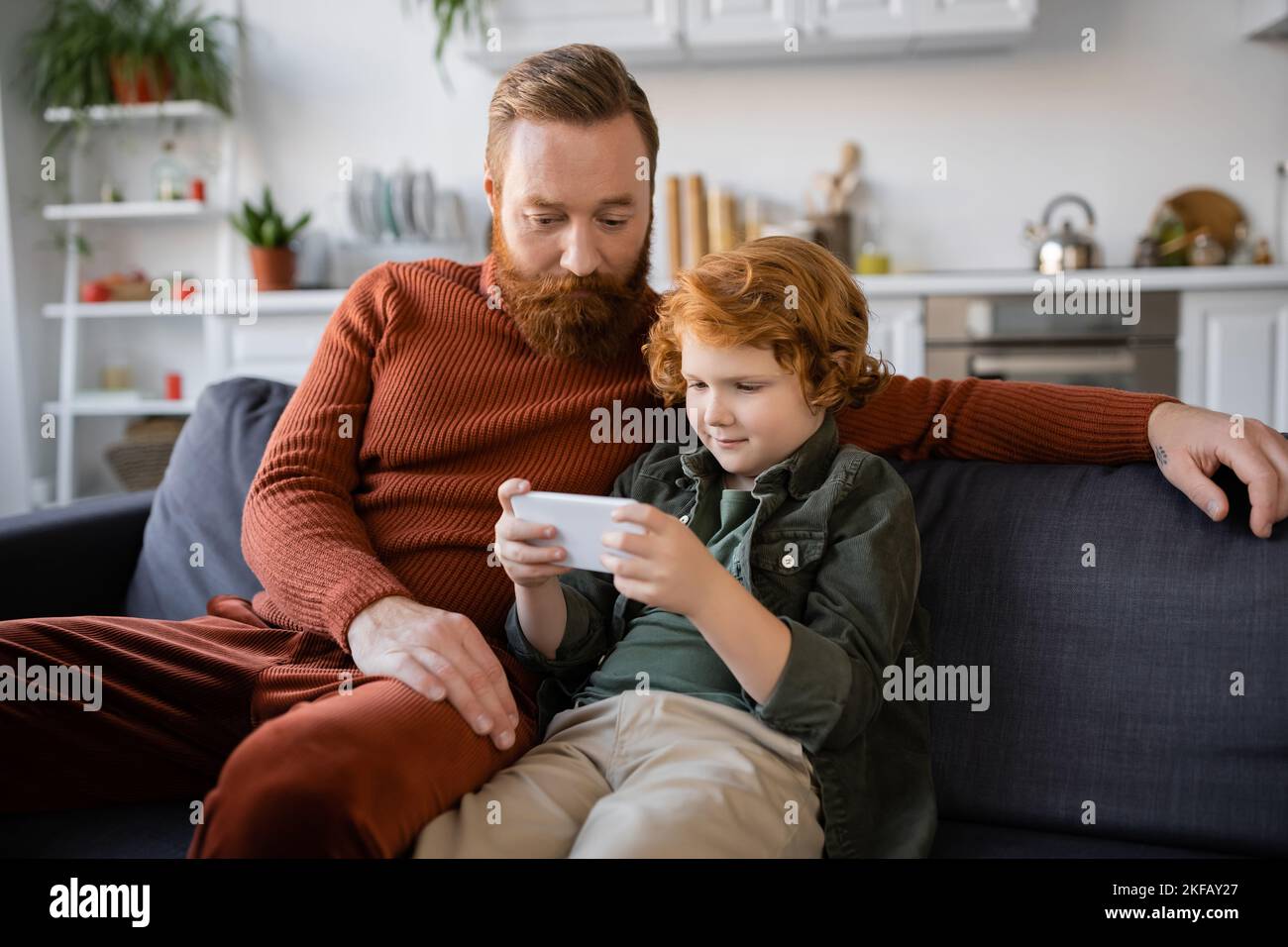 redhead boy sitting on couch near bearded dad and using smartphone ...