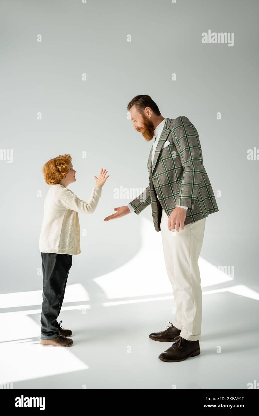 Side view of smiling redhead boy and father giving low five on grey ...