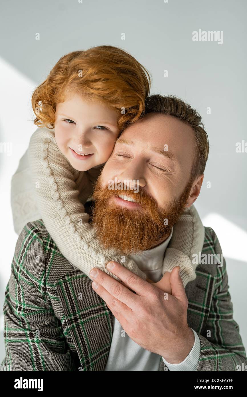 Positive redhead boy embracing bearded father in jacket on grey ...