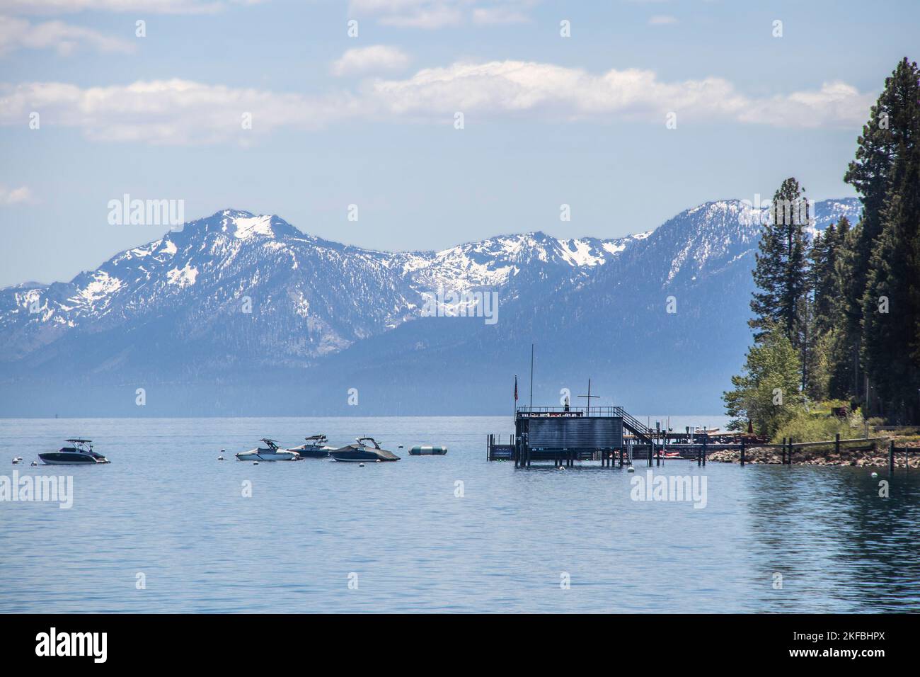 Lake Tahoe with boats moored and dock with American flag in foreground and snowy mountains in ...