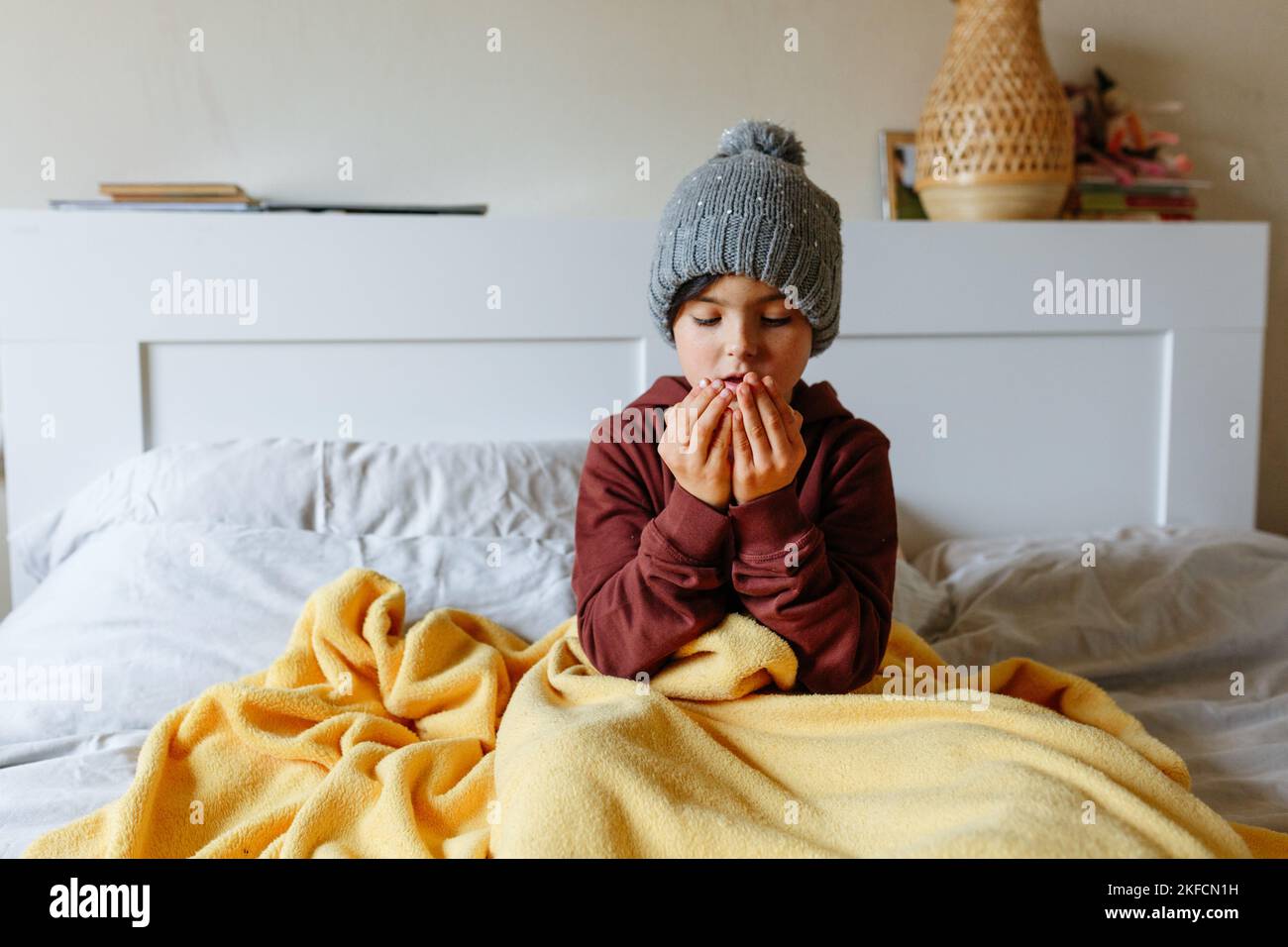 Child wearing grey warm hat sits on the bed under warm blanket and ...
