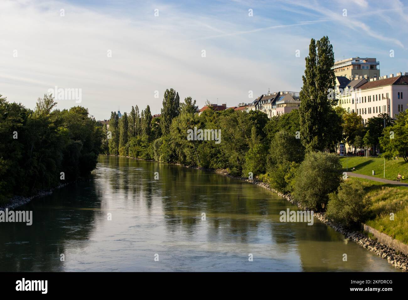 A scenic view of a tranquil river with green trees and buildings on its ...