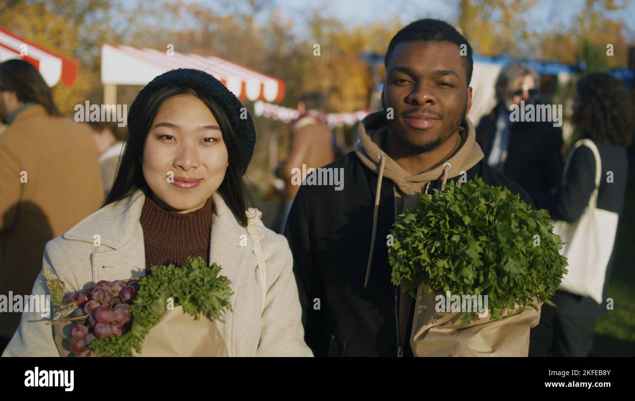 Diverse couple standing with paper bags of vegetables or fruits. Spouses looking at camera ...