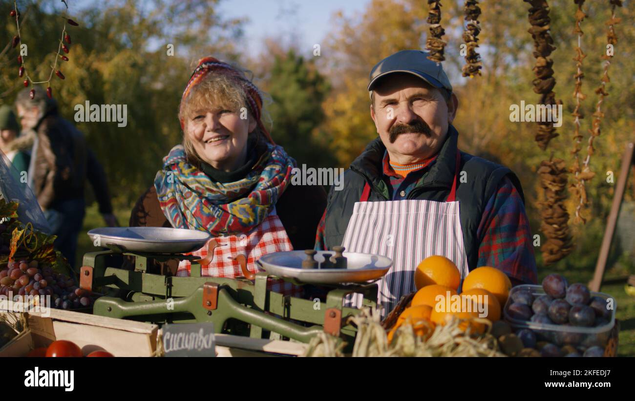 Happy elderly couple of farmers standing near fresh fruits and vegetables, looking at camera ...