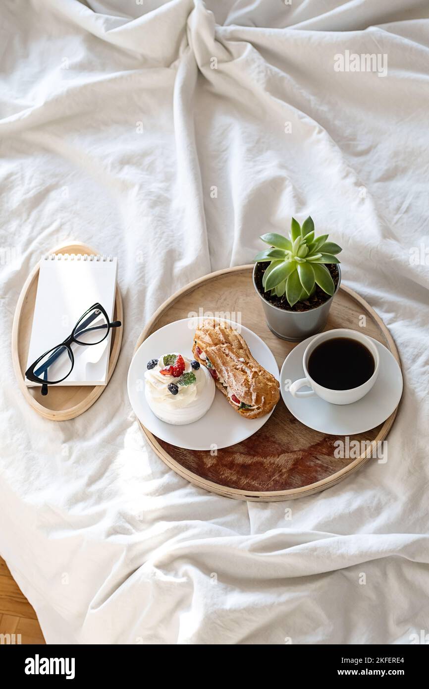 Cakes, coffee cup, notebook and eyeglass on wooden tray on bedding ...