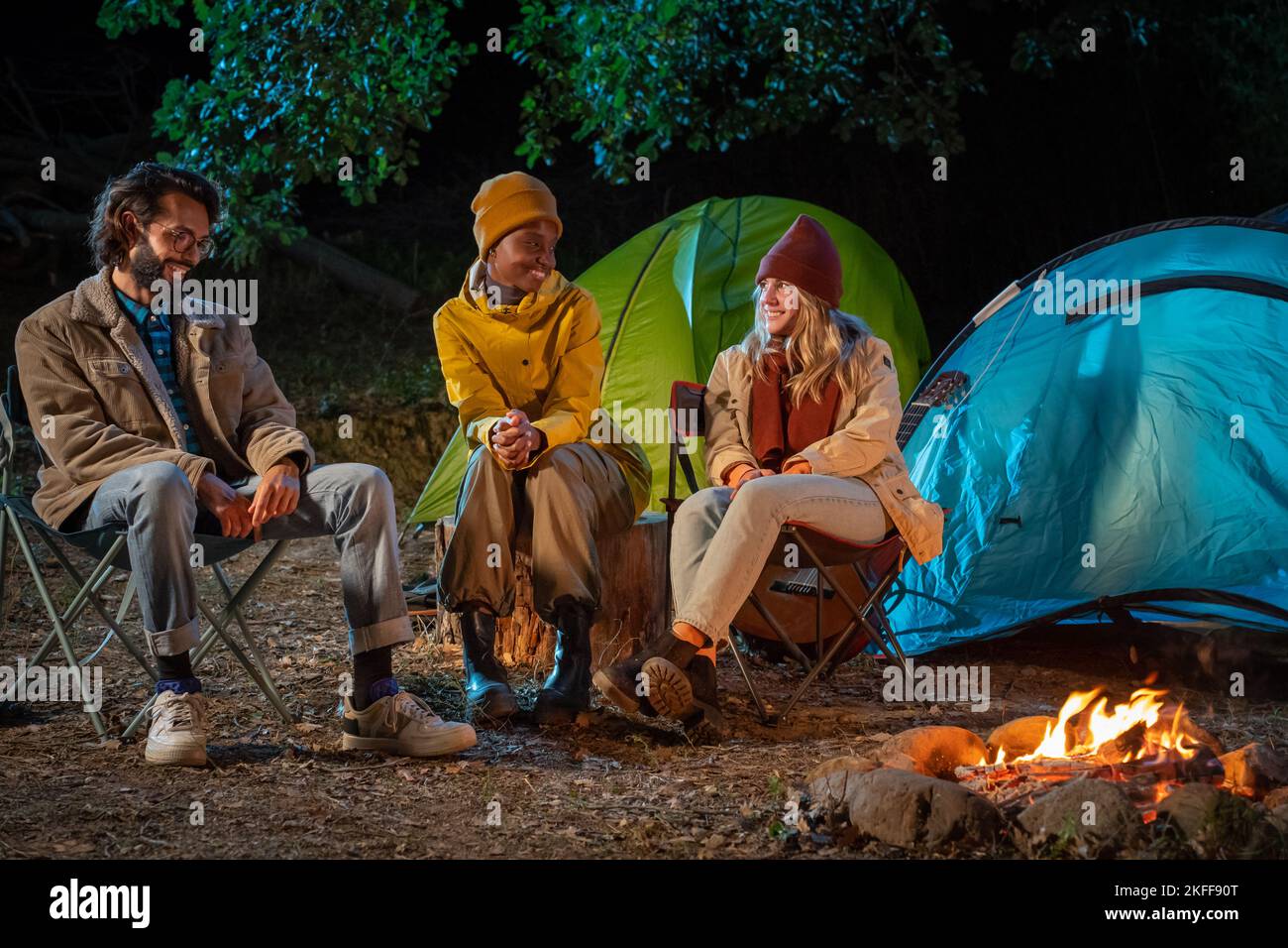 Three happy friends enjoying camping with tent chatting by a fire ...