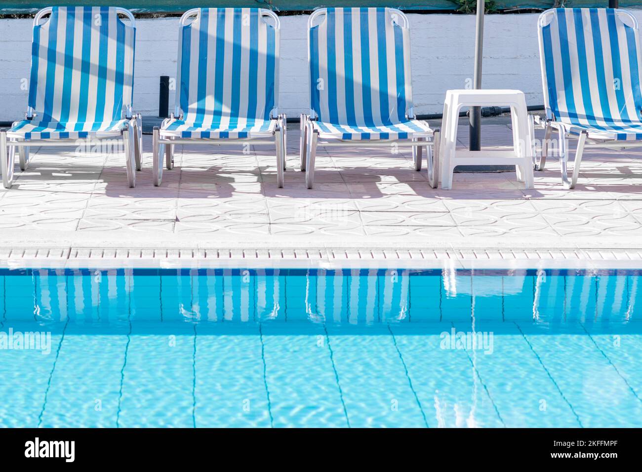 Retro blue and white sunbeds on the poolside of a swimming pool filled with clear blue water at ...