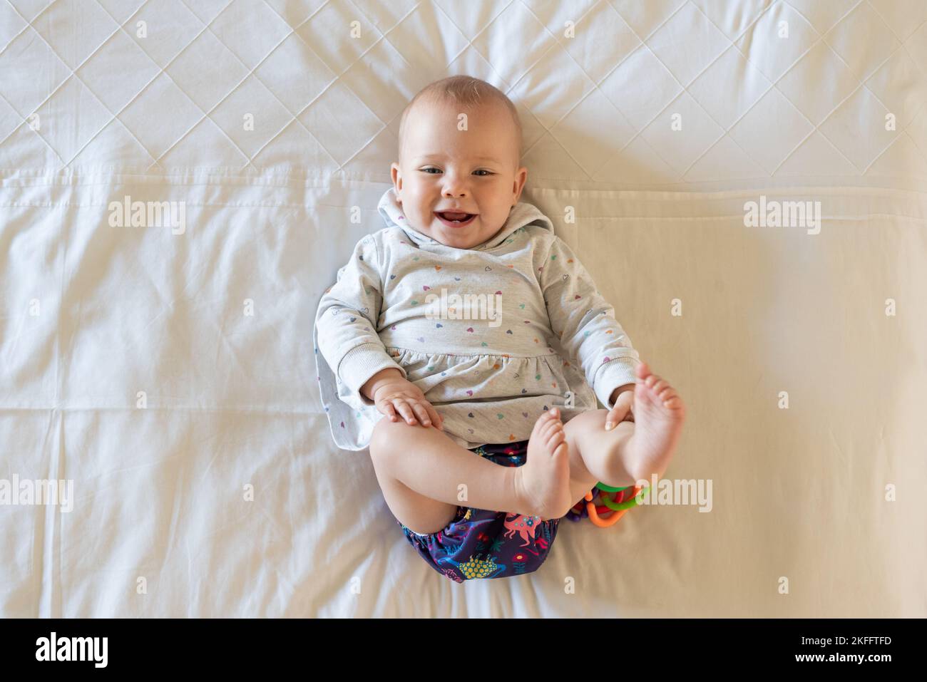Happy baby girl lying on her back on a bed. She is wearing a modern ...