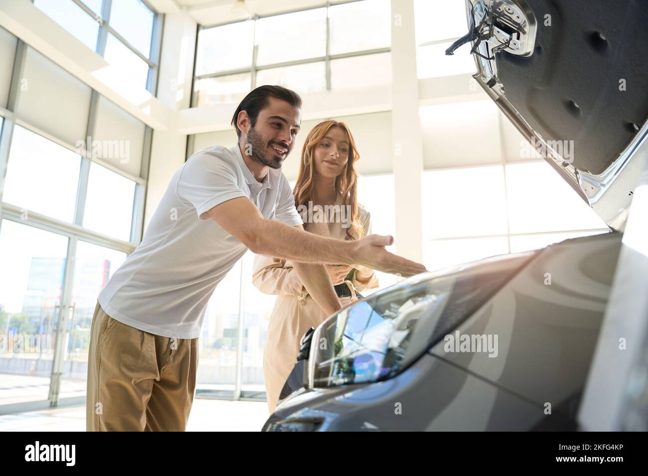 Pleased young man inspecting new motorcar in presence of saleswoman ...