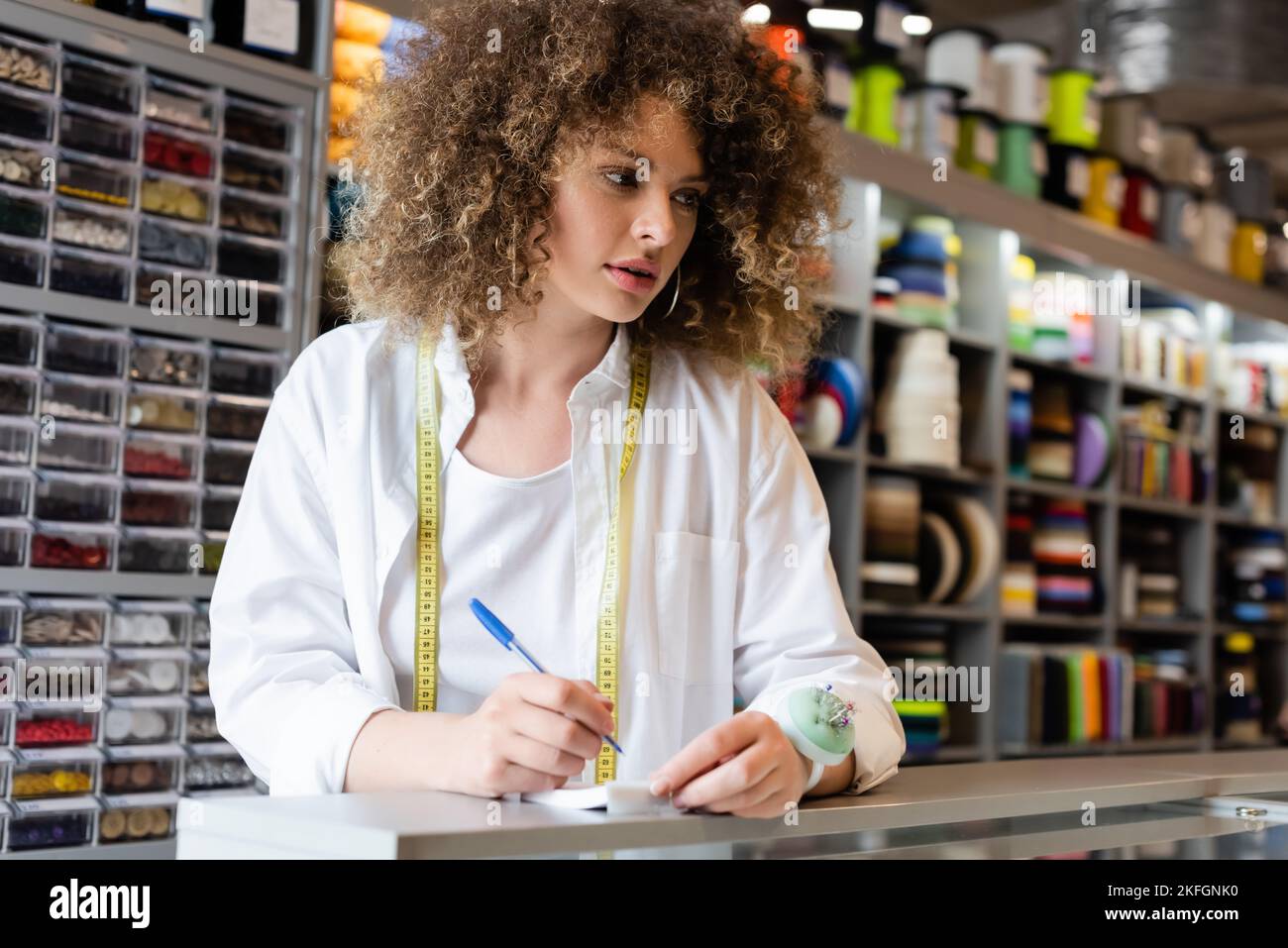 pretty and curly saleswoman making order near blurred rack with ...