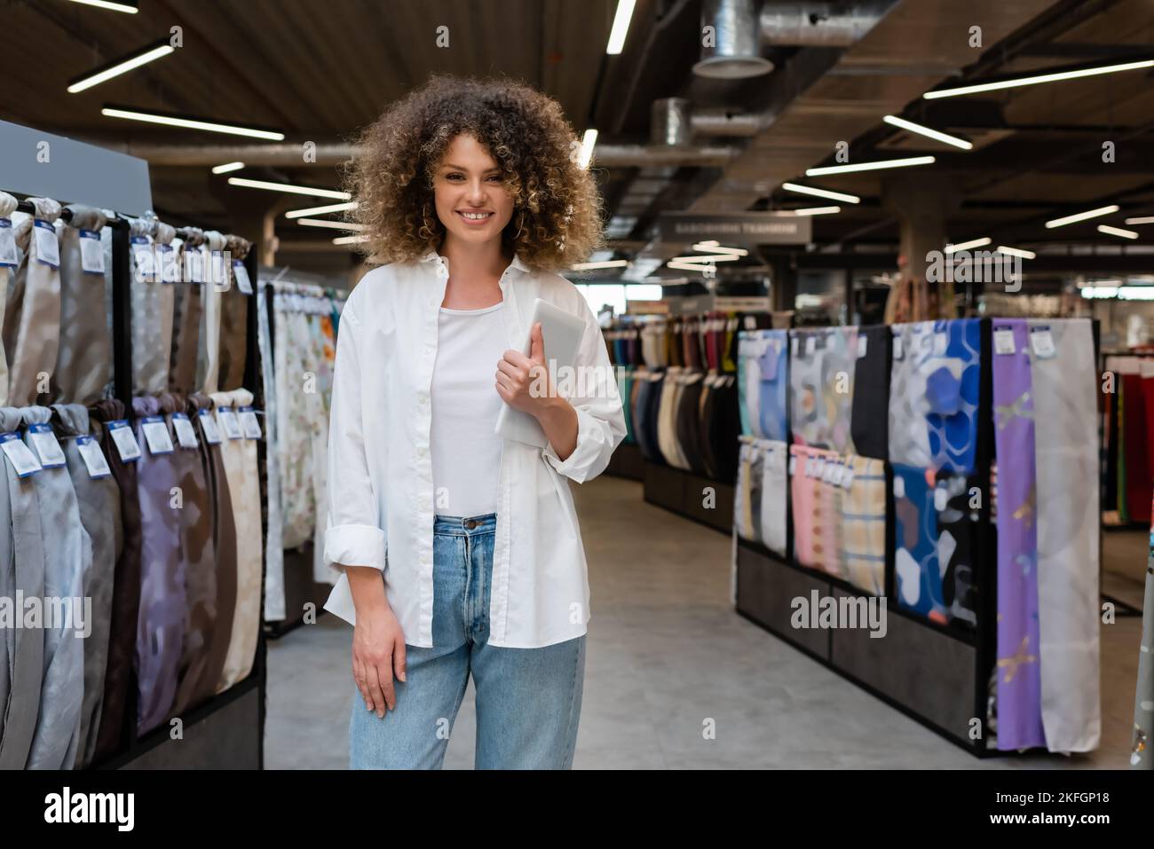 cheerful saleswoman holding digital tablet near different fabric in ...