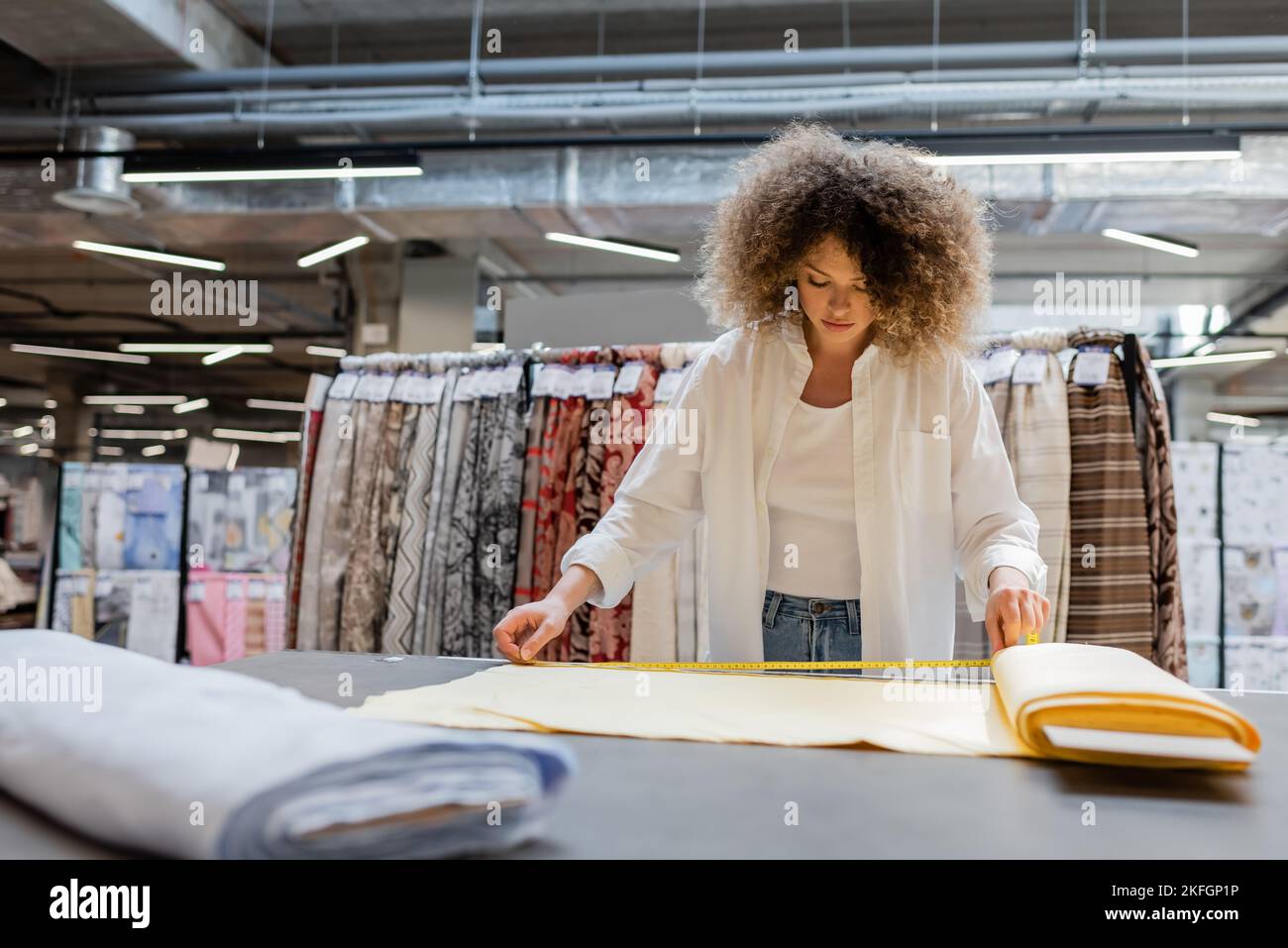 curly saleswoman measuring yellow fabric roll in textile shop Stock ...
