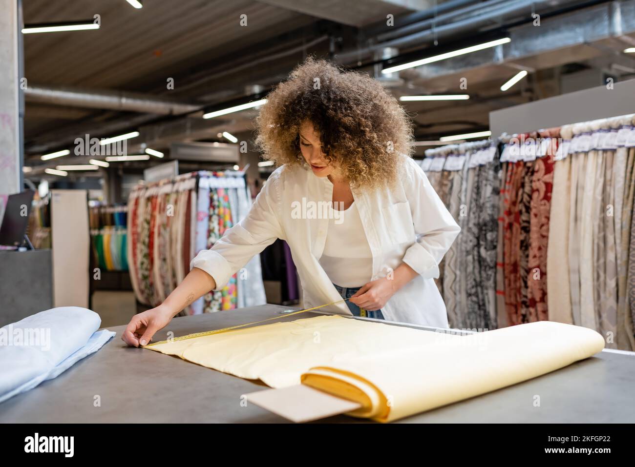 young and curly salesperson measuring yellow linen fabric roll in ...