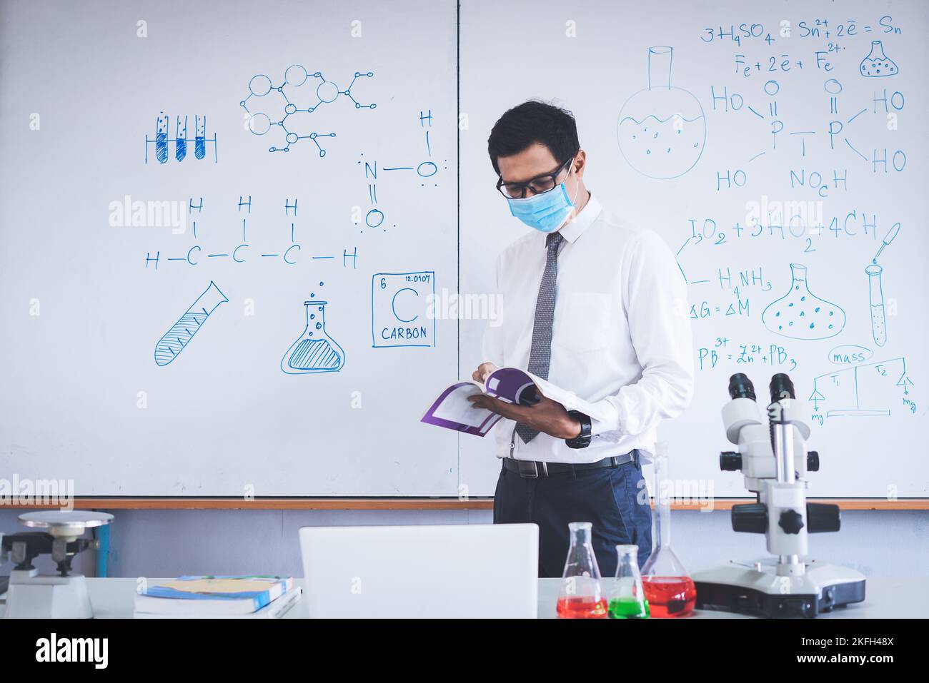 Science teacher wearing a mask stands looking at a textbook to teach ...