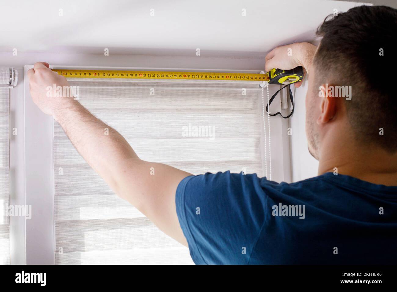 A handyman with a tape measure installs white roller blinds indoors ...