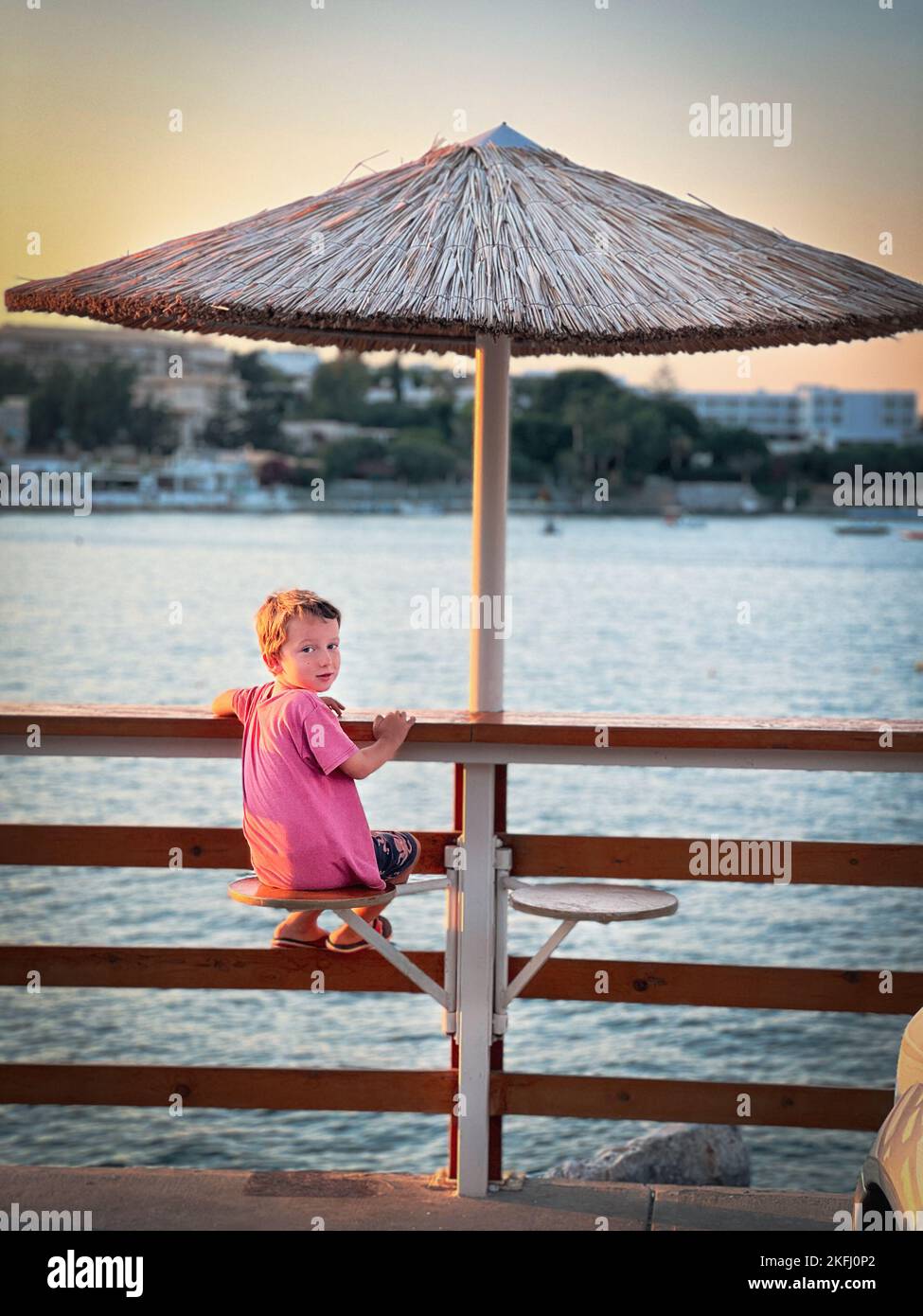 Portrait of cute boy sitting on railing under thatched roof in front of ...
