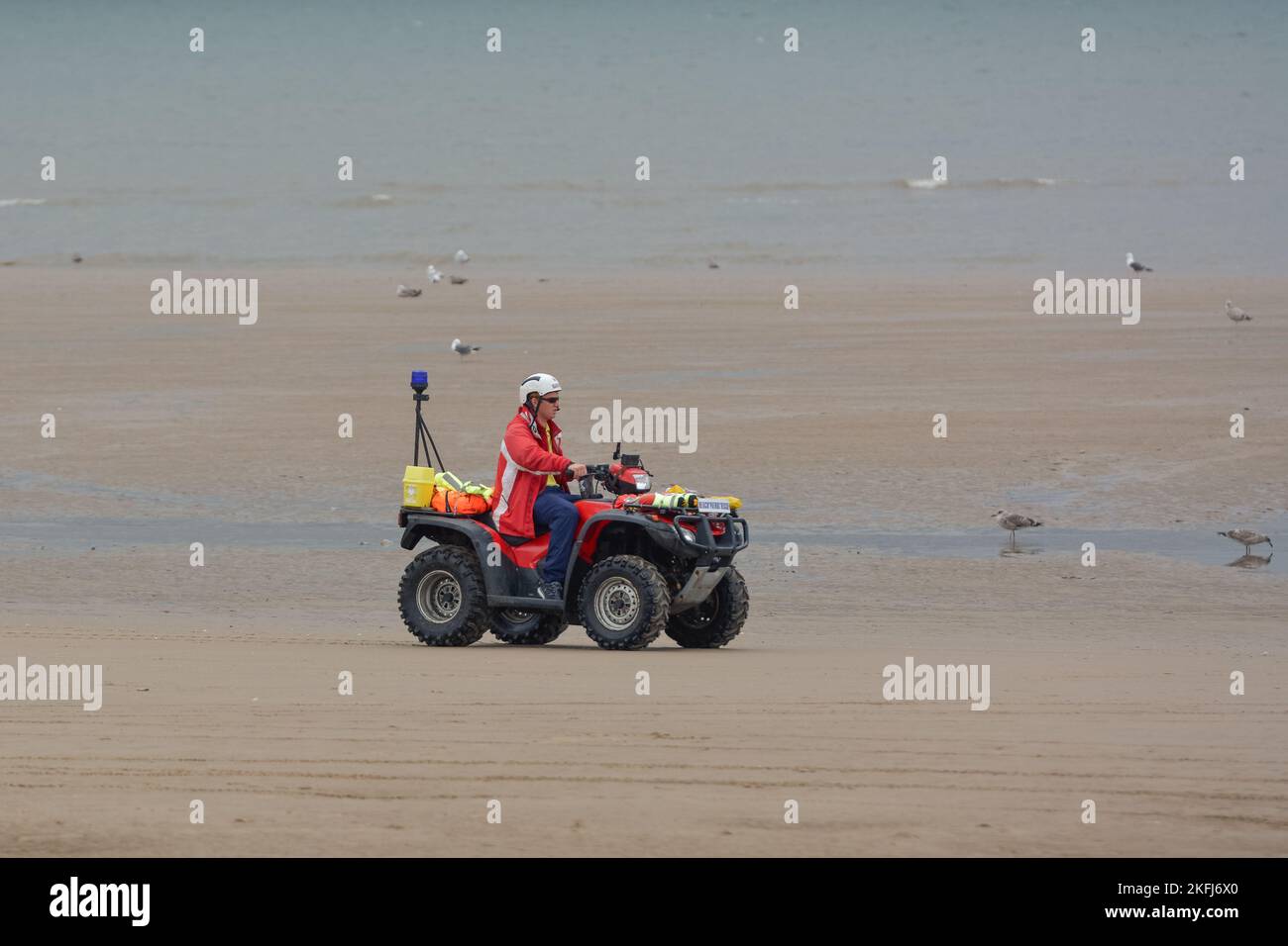RNLI Lifeboat man riding a quad bike along the beach. Red jacket and ...