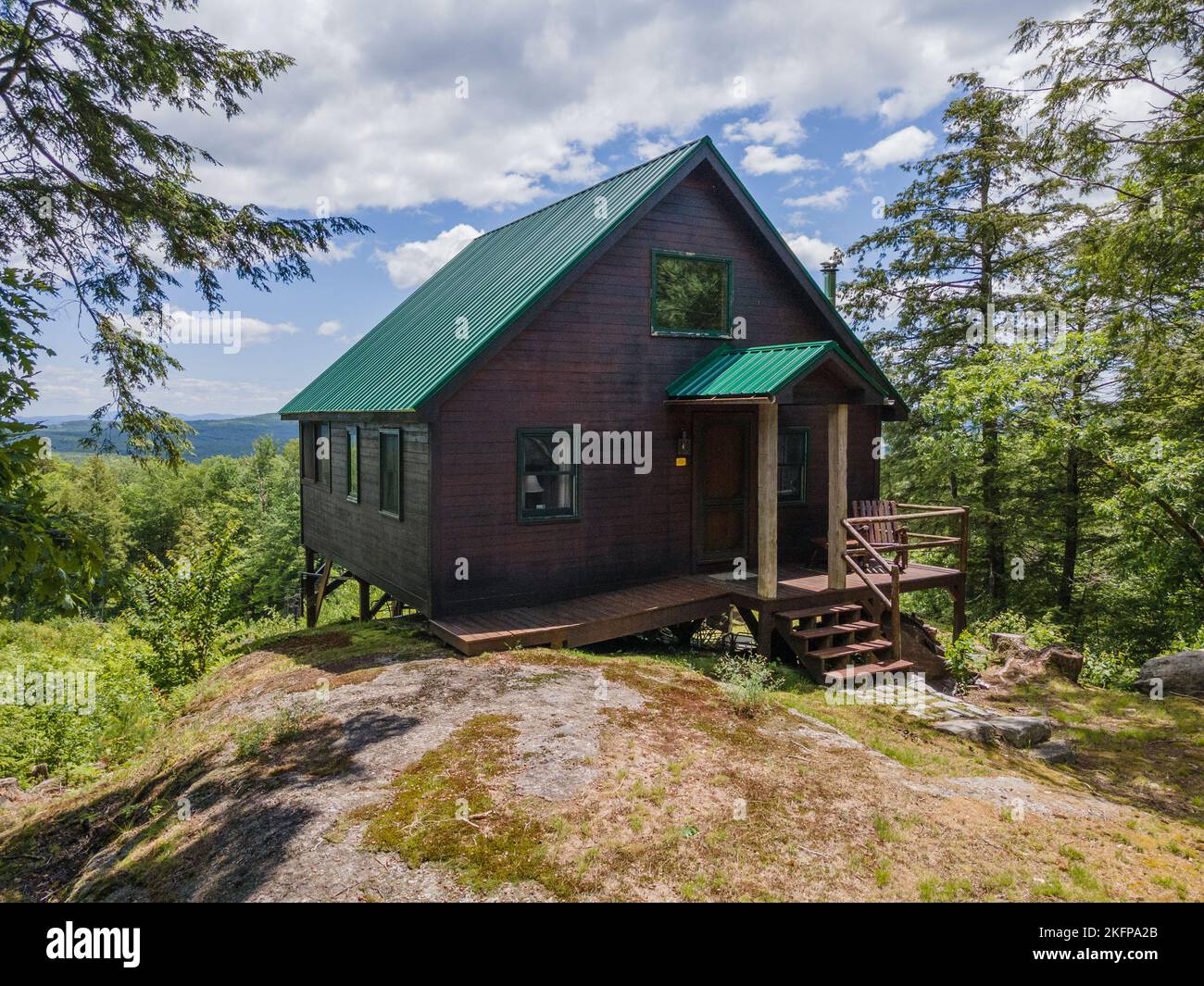A side view of a dark wooden cabin on a rock surrounded by greenery In ...